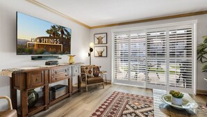 Uma sala de estar com televisão exibindo 'PALM SPRINGS', um console de madeira, um tapete estampado e uma porta de vidro deslizante com persianas.