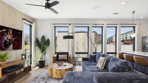 Uma sala de estar moderna com um sofá grande, uma mesa de centro, uma TV e janelas amplas com vista para a cidade.