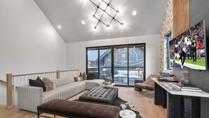 Uma sala de estar moderna com um sofá seccional, uma mesa de centro e um banquinho. Há uma janela grande com vista para uma casa e um pátio.