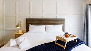 Cama bem arrumada com lençóis brancos, cabeceira de madeira, mesa de cabeceira com abajur e bandeja com muffin e xícara.