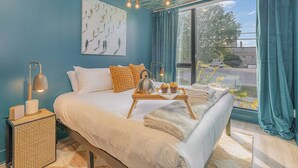 Quarto com cama, mesa de cabeceira, luminária e janela com cortinas.