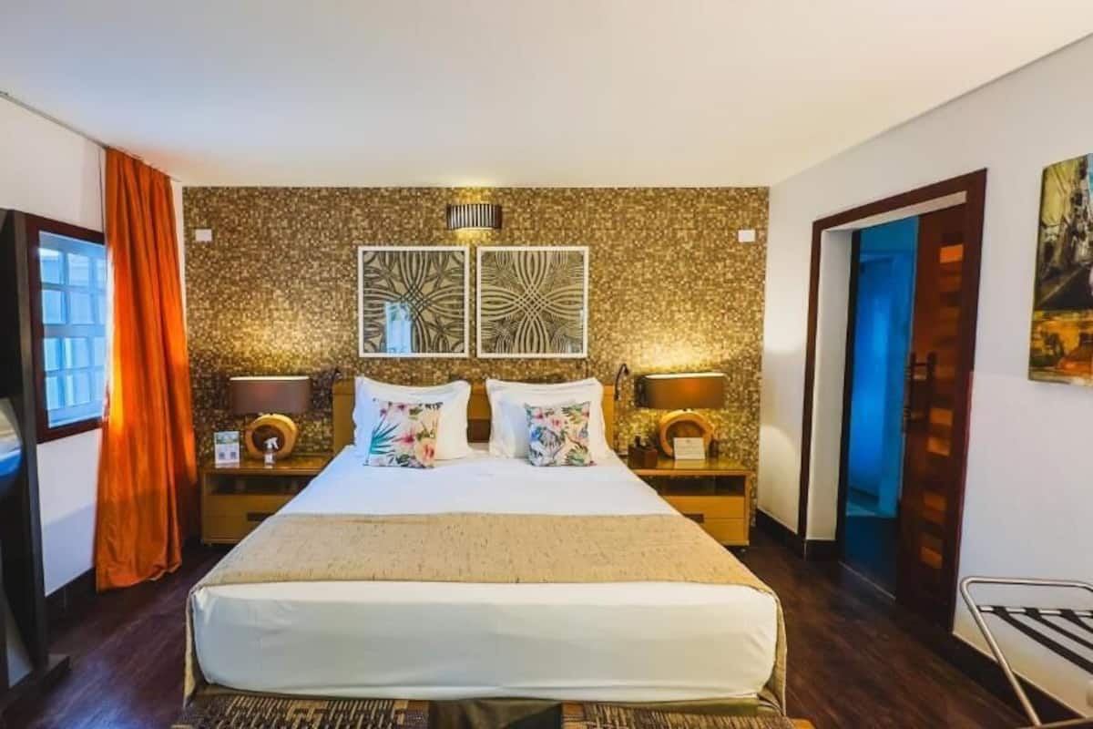 Quarto de hotel com uma cama grande, duas mesas de cabeceira, uma luminária na parede e uma janela com cortinas.