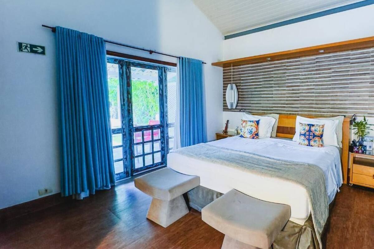 Quarto com cama, um banco, janela com cortinas e vista para o verde.