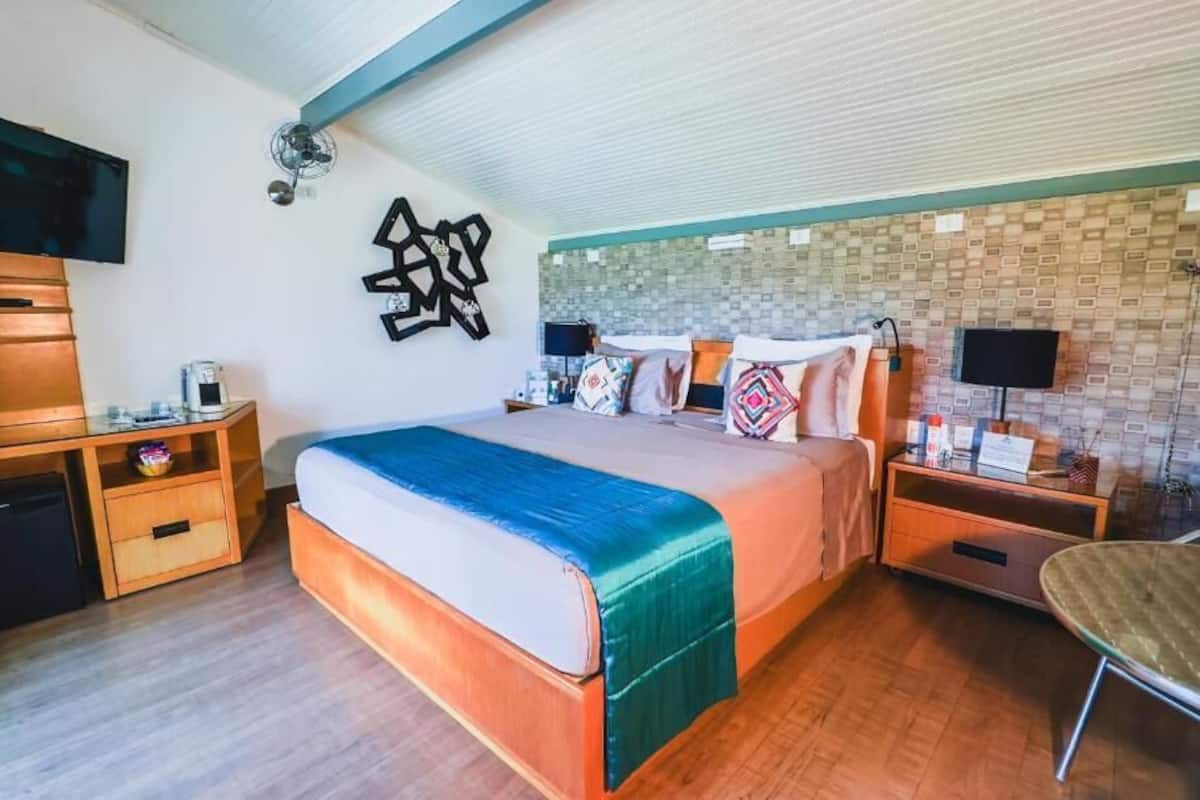 Quarto de hotel moderno com uma cama grande, mesas de cabeceira, televisão e uma pequena área de trabalho.