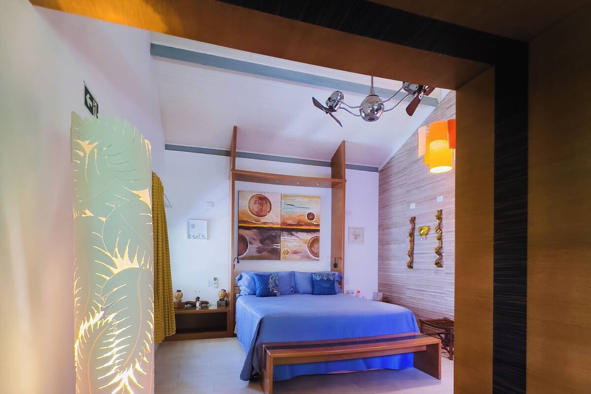 Quarto com cama, mesas de cabeceira, ventoinha no teto e luminária na parede.