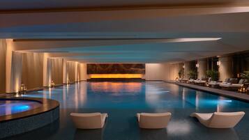Uma piscina interior ampla com hidromassagem, espreguiçadeiras e uma parede de mármore ao fundo.