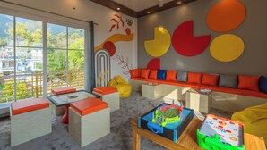 Uma área de recreação infantil com mobiliário colorido, uma grande janela com vista para vegetação e uma parede com arte abstrata.
