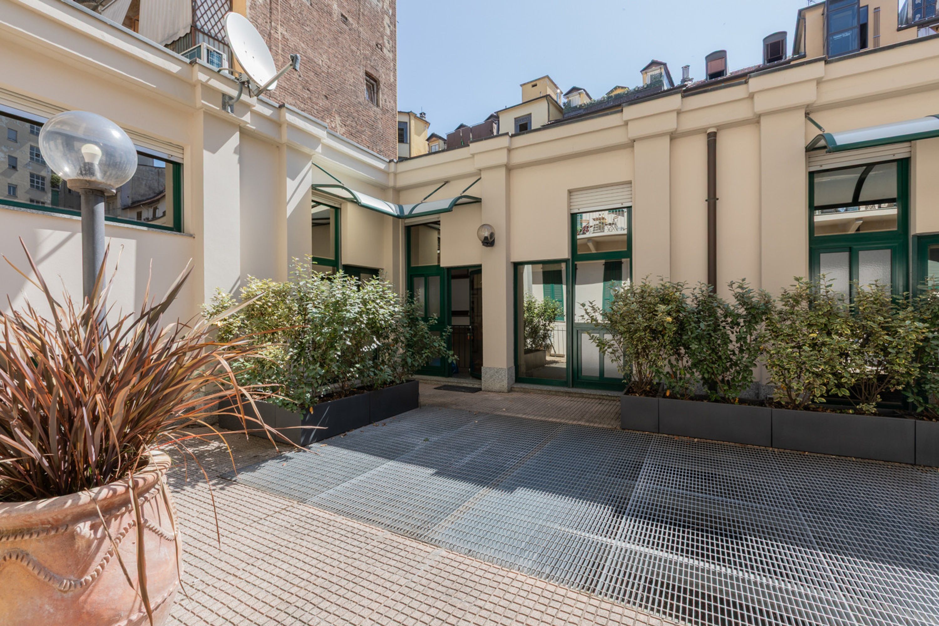 Um pátio com piso de azulejos, plantas em vasos e um edifício com portas e janelas verdes.