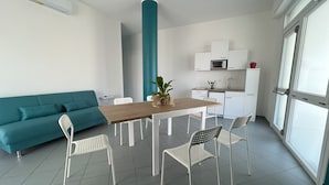 Um espaço de estar moderno com um sofá azul-esverdeado, uma mesa de jantar de madeira, cadeiras brancas, uma área de cozinha com armários brancos e uma grande janela.