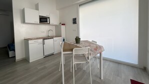Uma cozinha compacta com armários brancos, micro-ondas e pia. Há uma mesa de jantar com cadeiras e uma planta em vaso.
