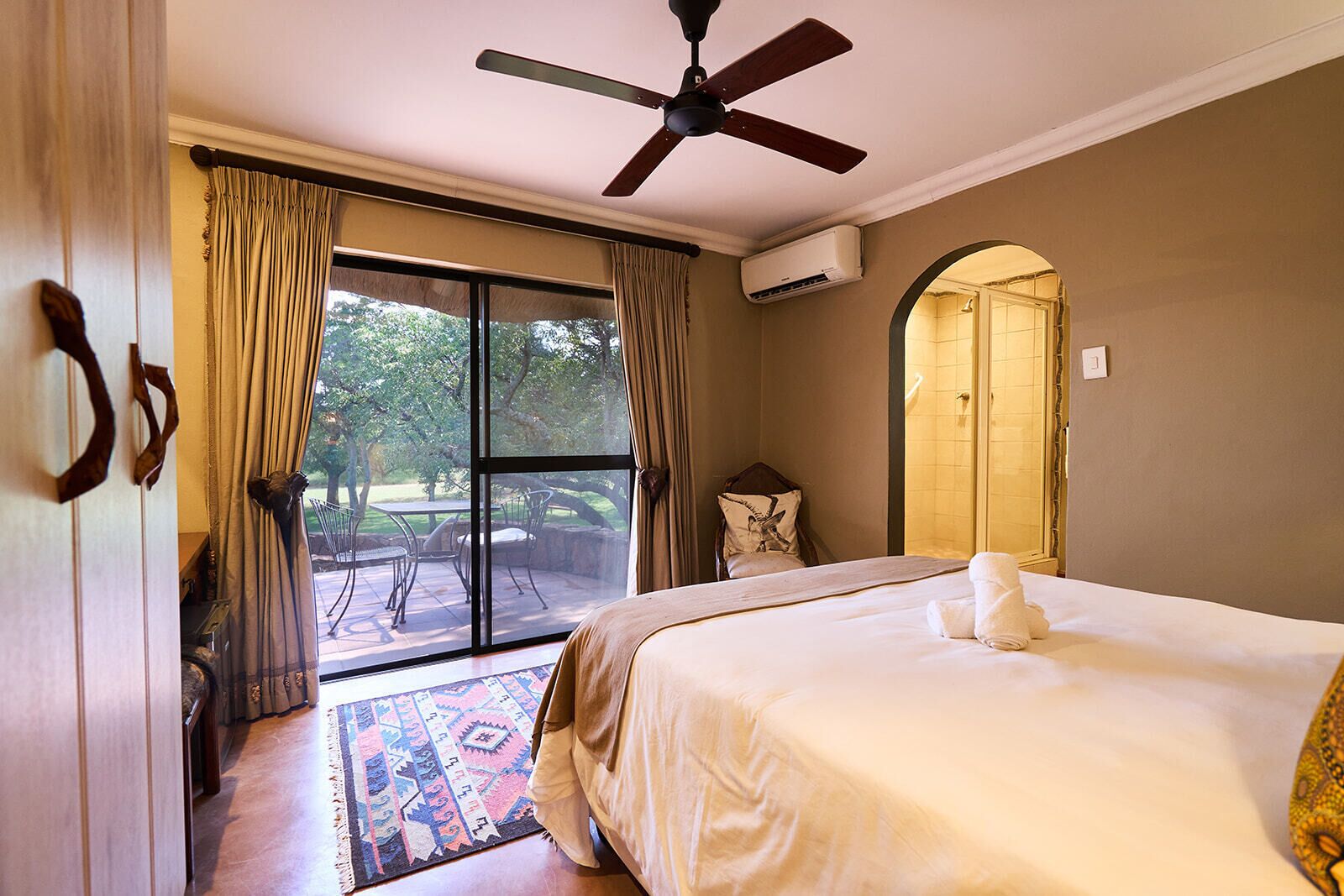 Quarto de hotel com cama, ventoinha no teto, janela grande com cortinas, mesa pequena com cadeiras e ar condicionado.