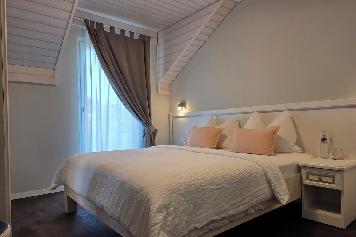 Quarto com cama grande, mesa de cabeceira, janela com cortinas e teto inclinado.