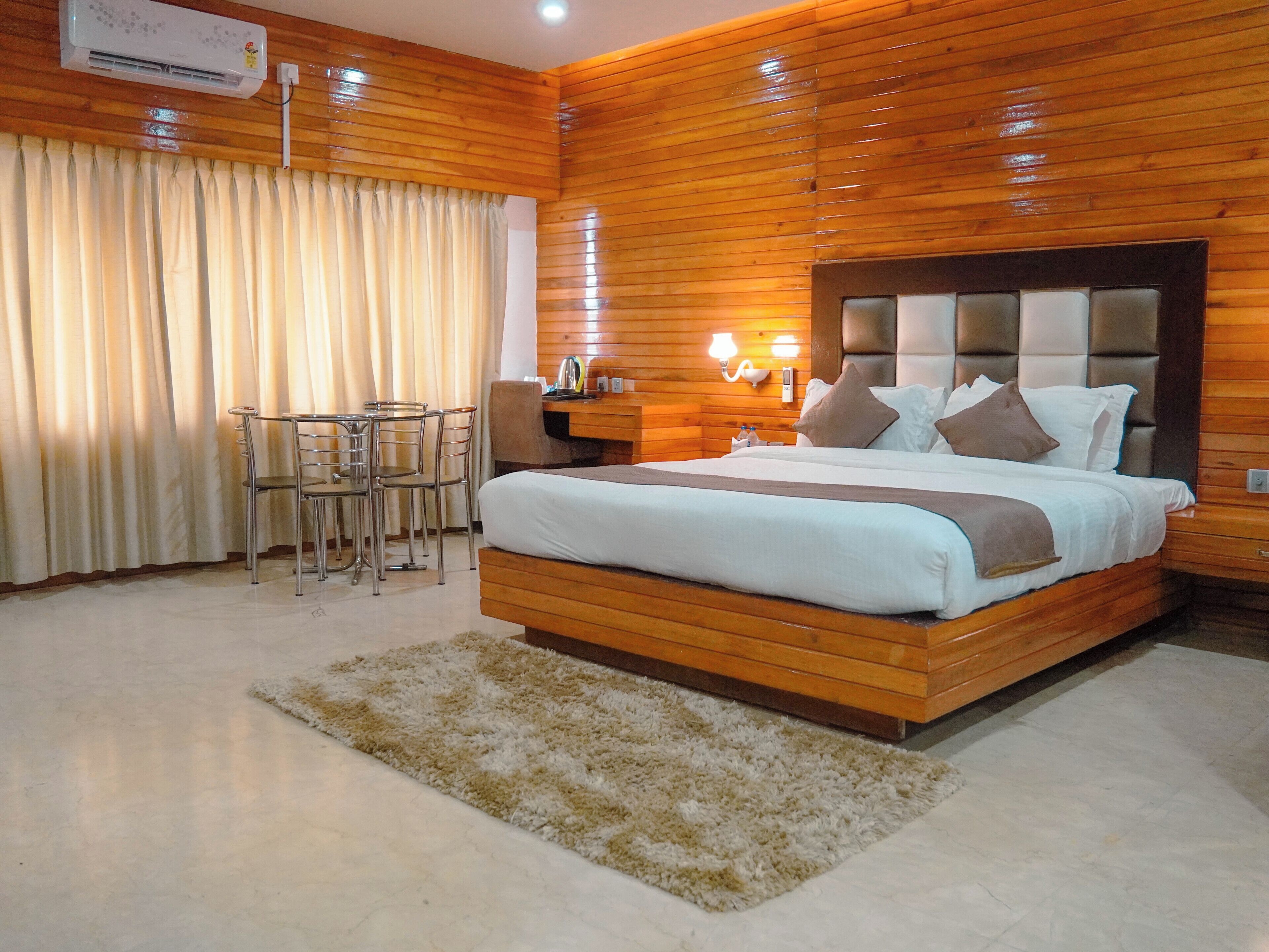 Quarto de hotel com parede de painéis de madeira, cama com roupa de cama branca, mesa pequena com cadeiras e janela com cortinas.