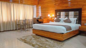 Quarto de hotel com parede de painéis de madeira, cama com roupa de cama branca, mesa pequena com cadeiras e janela com cortinas.