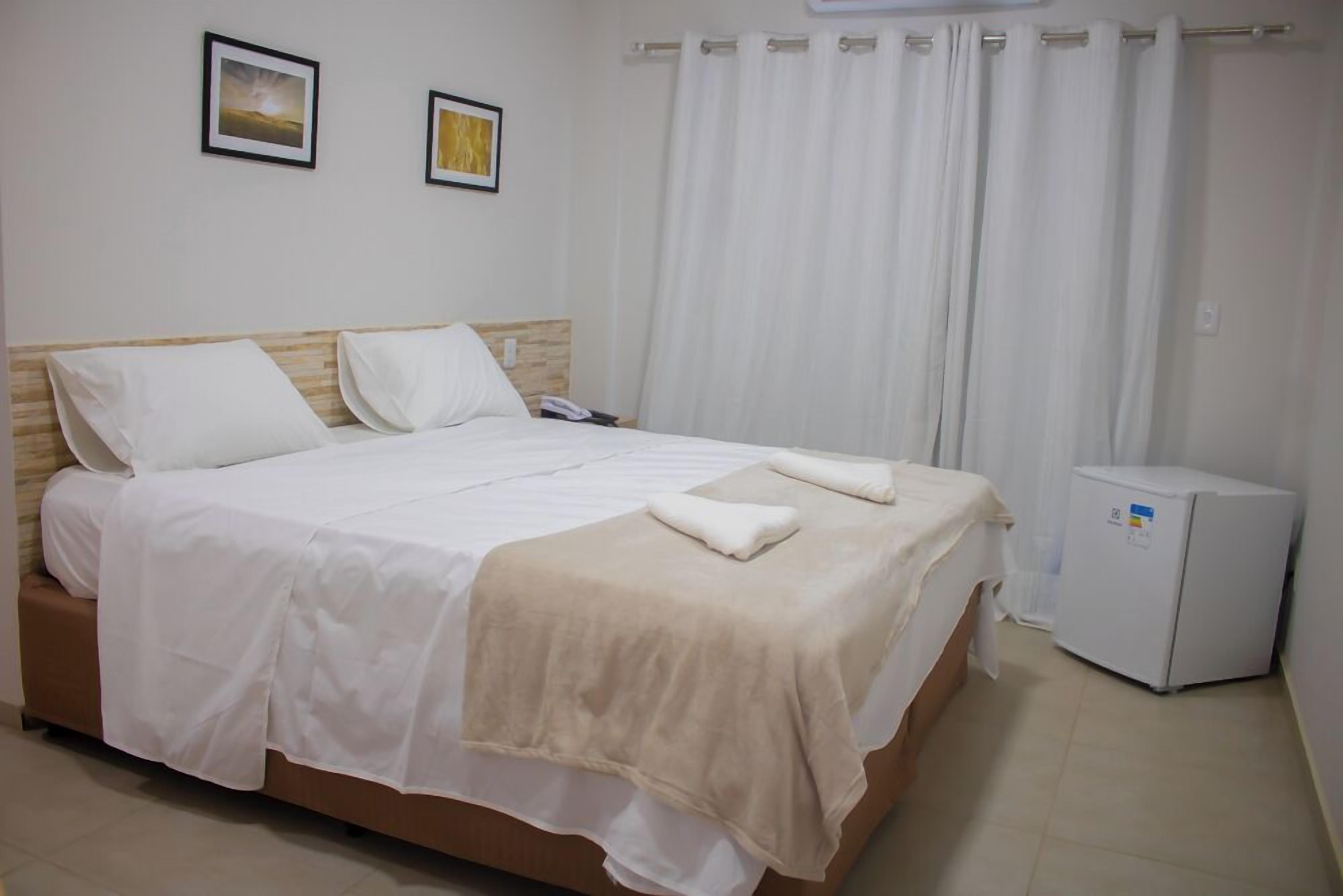 Cama bem arrumada com lençóis brancos e manta bege, mesinha de cabeceira com telefone, e refrigerador branco em quarto de hotel.
