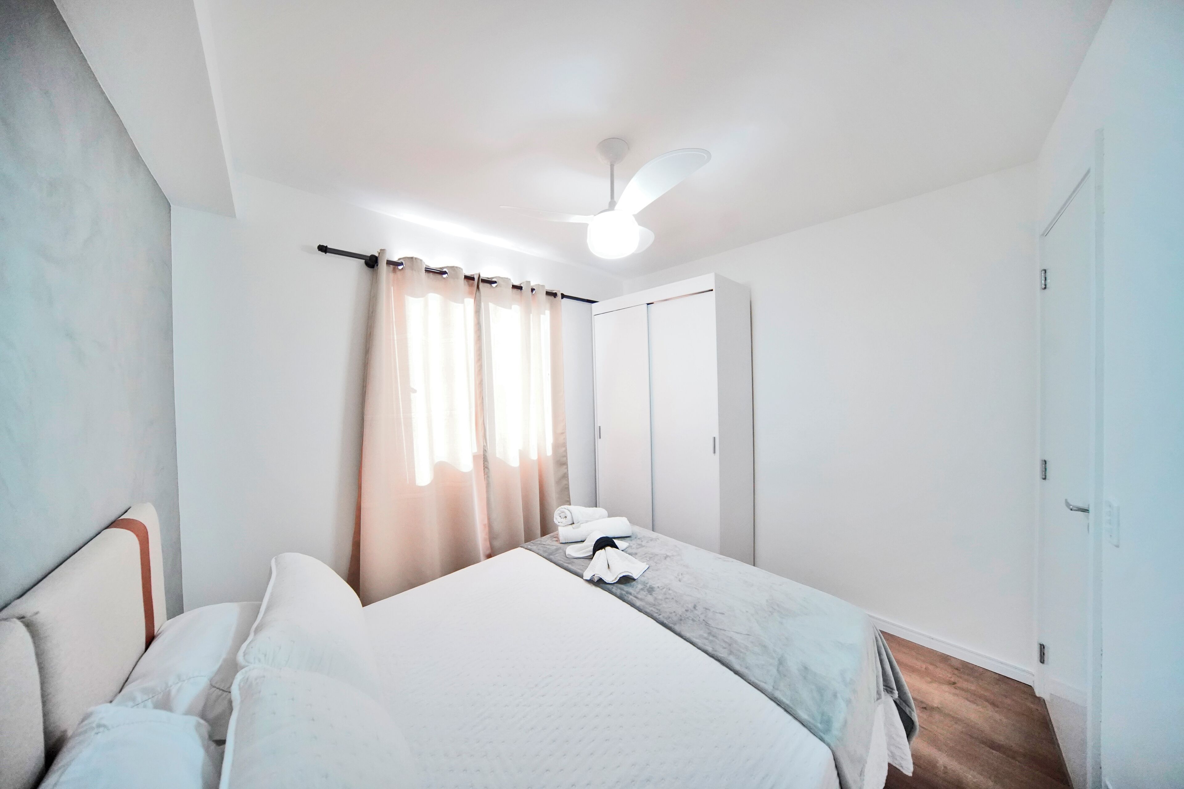 Quarto com cama, ventoinha no teto, guarda-roupa e janela com cortinas.