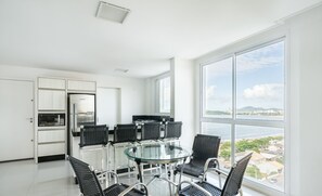 Cozinha moderna com armários brancos e eletrodomésticos de aço inoxidável, mesa de jantar de vidro com cadeiras pretas e uma grande janela com vista para o mar e a paisagem urbana.