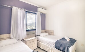 Quarto com duas camas, janela com cortinas, ar condicionado e uma pequena mesa de cabeceira.