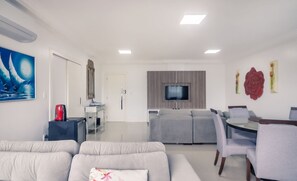 Uma sala de estar moderna com um sofá cinzento, uma mesa de jantar com cadeiras, uma televisão de tela plana na parede e uma área de cozinha ao fundo.