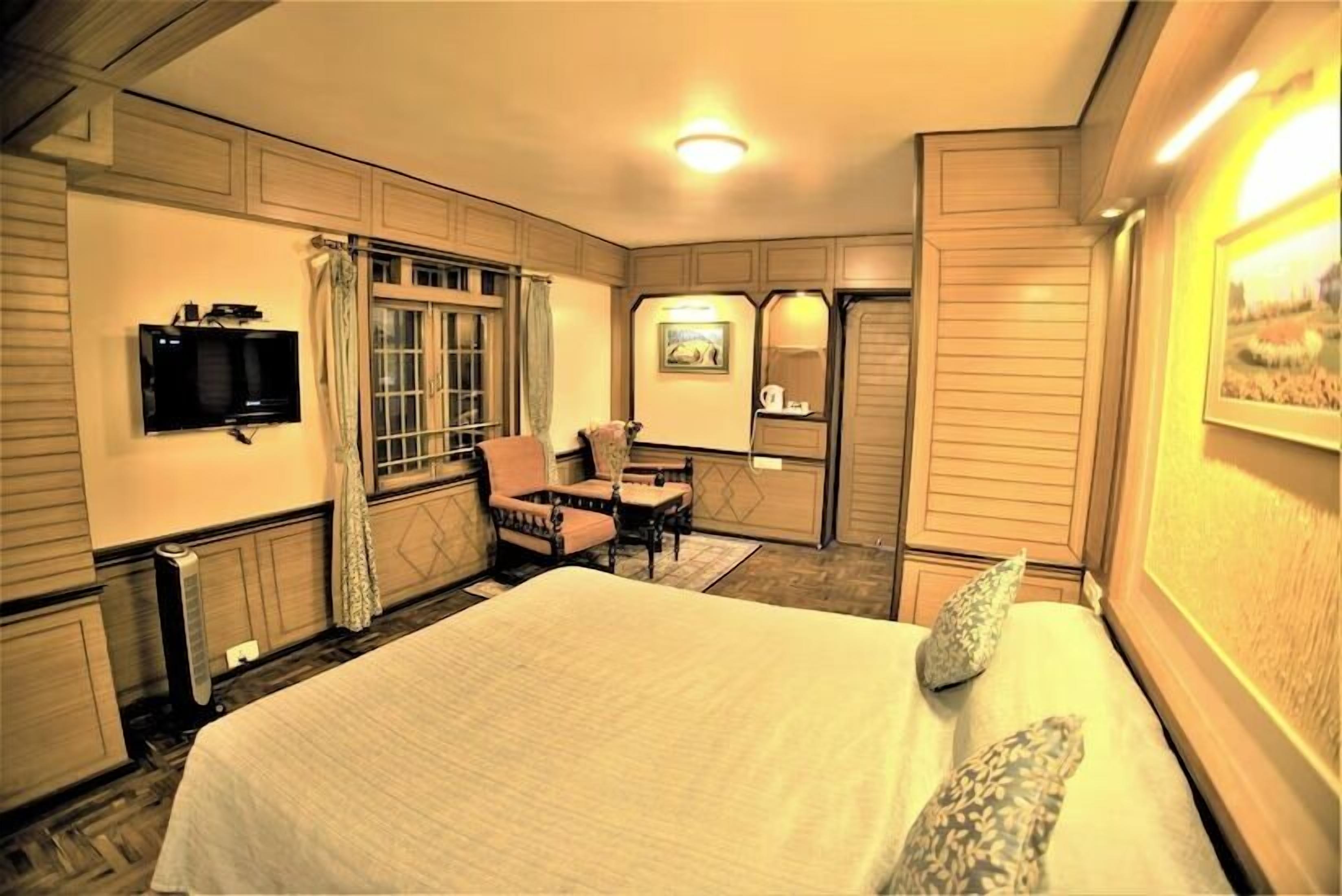 Quarto de hotel com uma cama grande, televisão na parede, uma cadeira, uma mesinha e uma janela com cortinas.