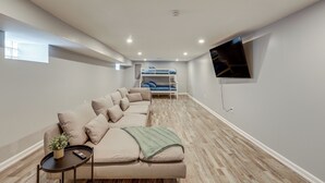 Uma sala de estar moderna com um sofá seccional, uma mesa pequena com uma planta, uma televisão na parede e uma cama beliche ao fundo.