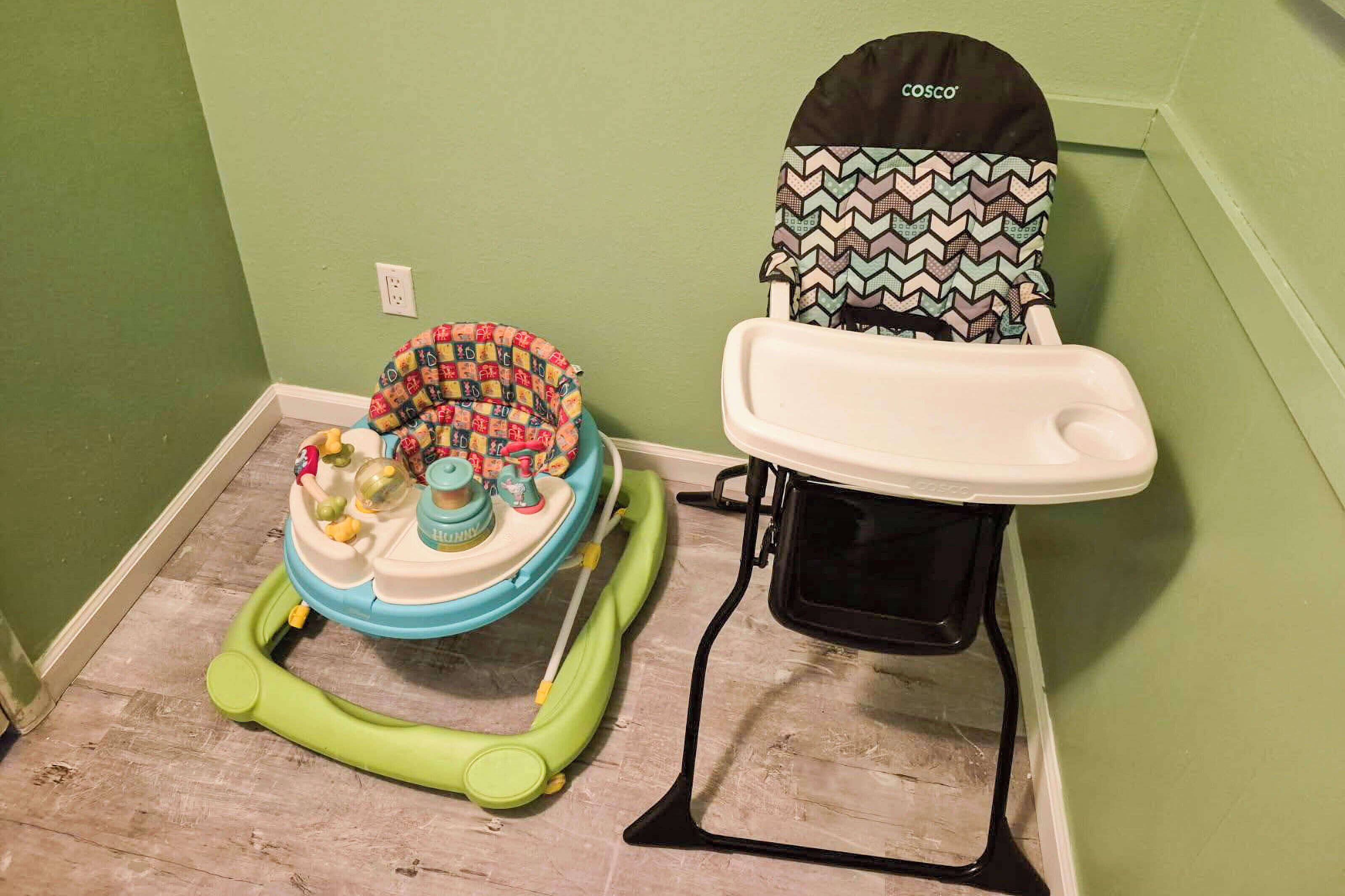 Quarto com parede verde, um andador para bebê e uma cadeirinha de alimentação.