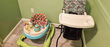 Quarto com parede verde, um andador para bebê e uma cadeirinha de alimentação.
