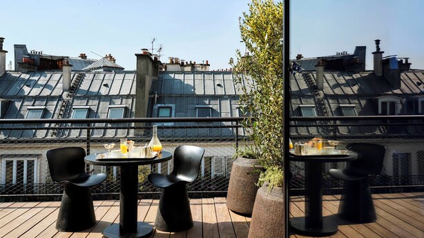 Terraço no telhado com mesas e cadeiras, uma planta em vaso e vista para edifícios com telhados inclinados.