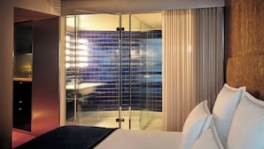 Quarto de hotel com uma cama, televisão, porta de vidro que dá para uma varanda e vista para o horizonte urbano.
