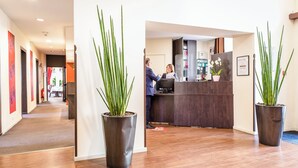 Uma área de recepção com piso de madeira, dois vasos grandes com plantas e um balcão de atendimento.