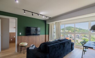Uma sala de estar moderna com uma parede verde decorativa, uma TV de tela plana, um sofá escuro, uma mesinha redonda com livros e vista para a paisagem urbana através de amplas janelas.