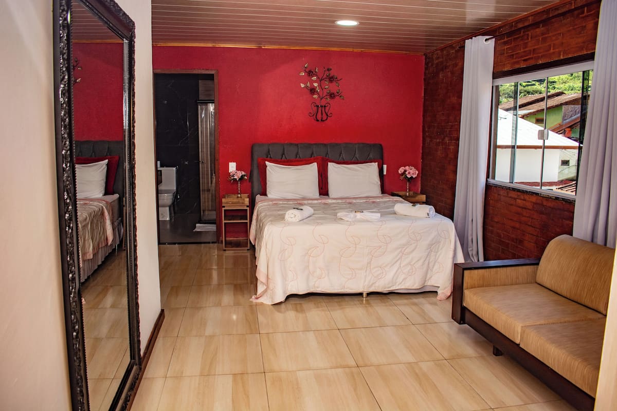 Quarto de hotel com uma cama grande, um sofá, um espelho e vista para o exterior.