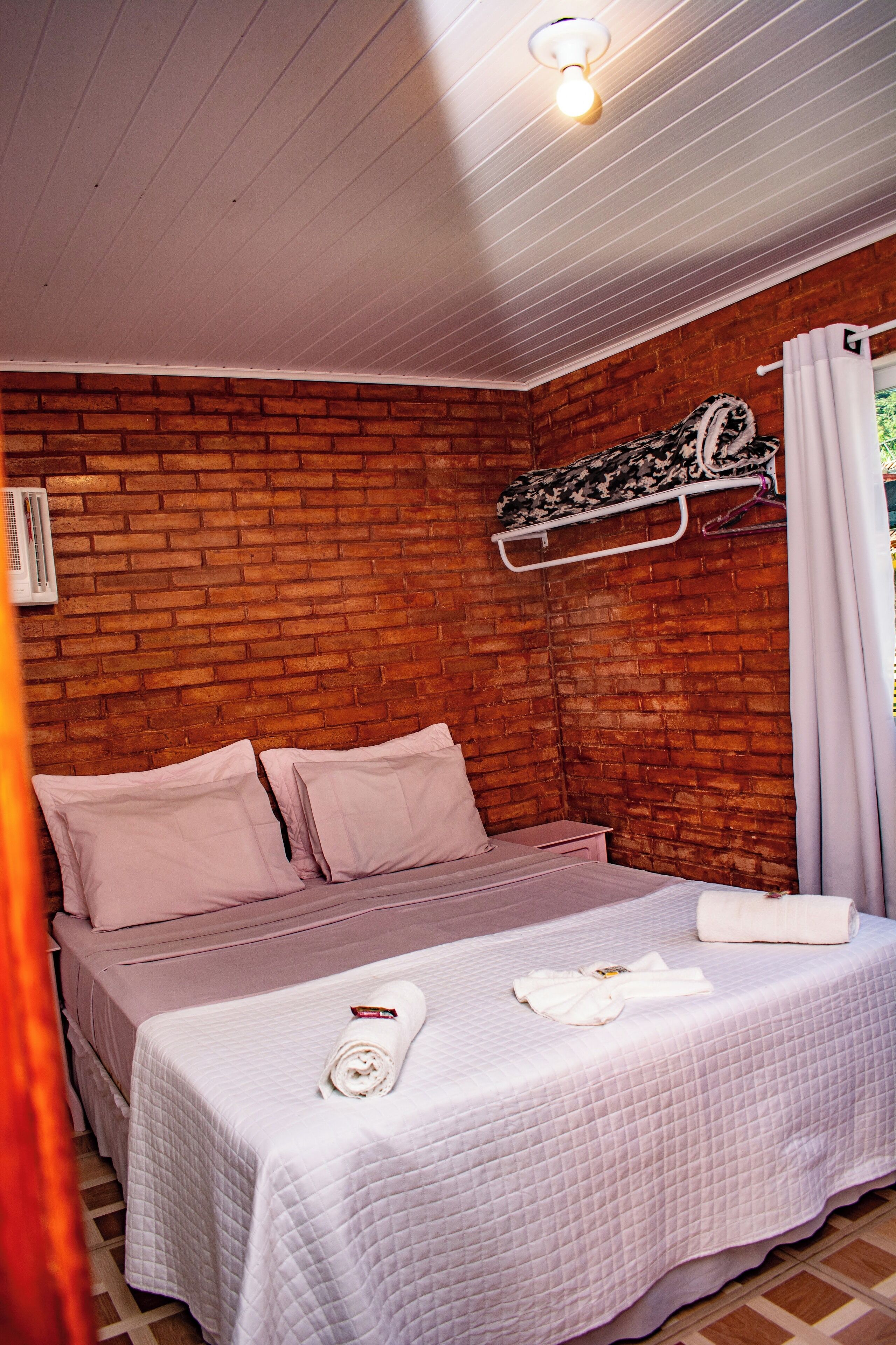 Quarto com parede de tijolos, cama com colcha acolchoada branca, varal de roupa e janela com cortinas.