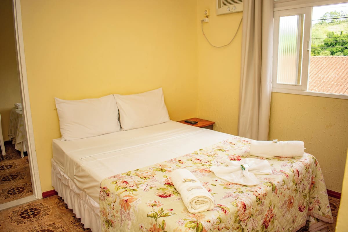 Uma cama bem arrumada com roupa de cama floral e uma toalha enrolada, uma mesa de cabeceira com controle remoto e uma janela com vista para o verde.