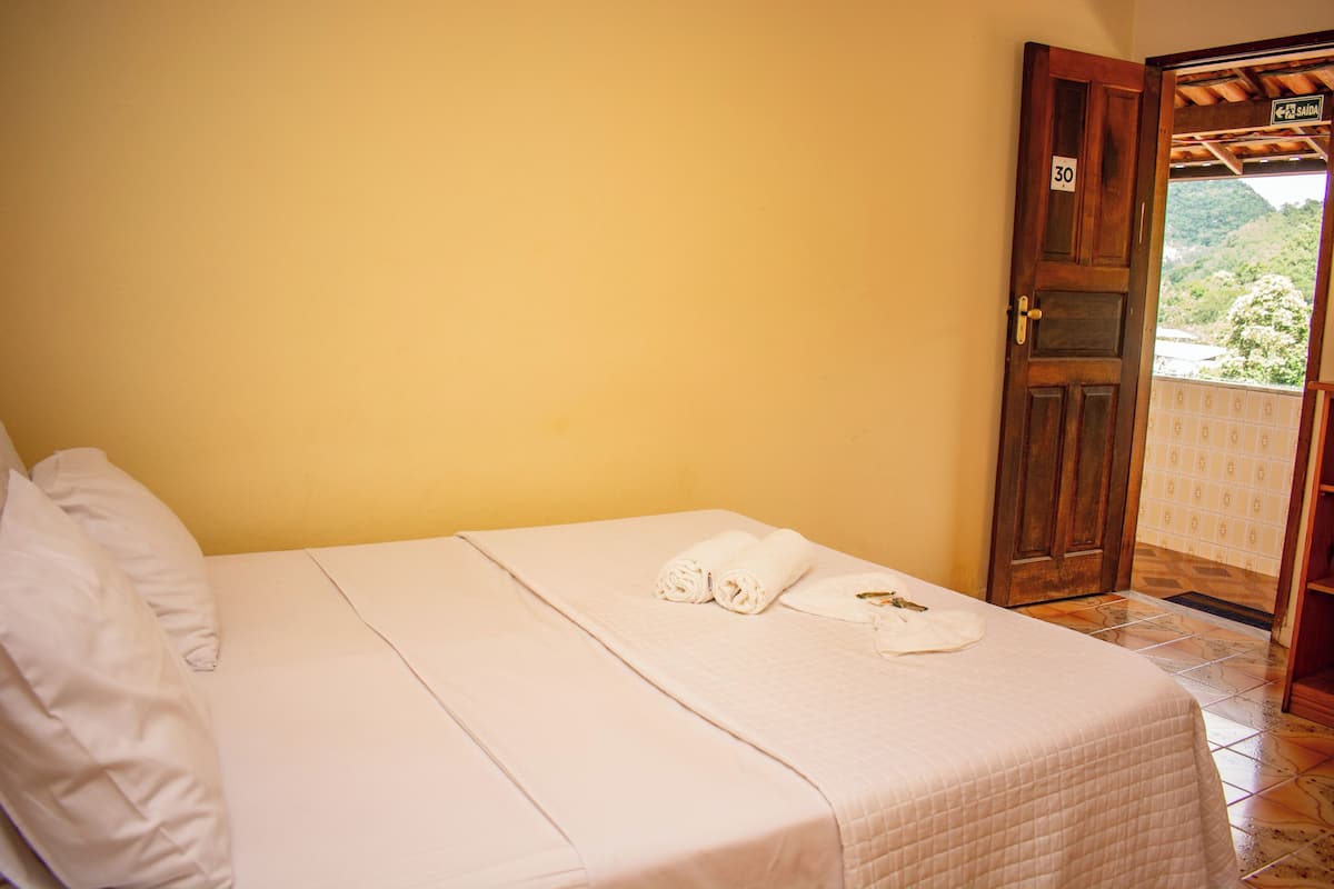 Cama bem arrumada com lençóis brancos e uma toalha dobrada em quarto com paredes amarelas e porta de madeira.