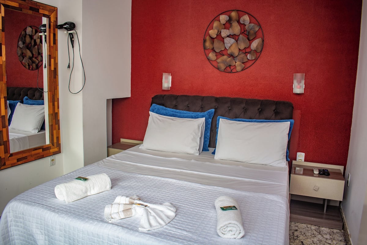 Quarto de hotel com cama, mesa de cabeceira, espelho, um enfeite na parede e um secador de cabelo fixado na parede.