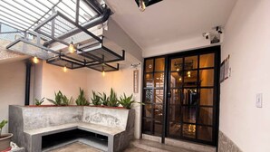 Um interior moderno com um banco de concreto, plantas em vasos, entrada por uma porta de vidro e estrutura de teto em metal.
