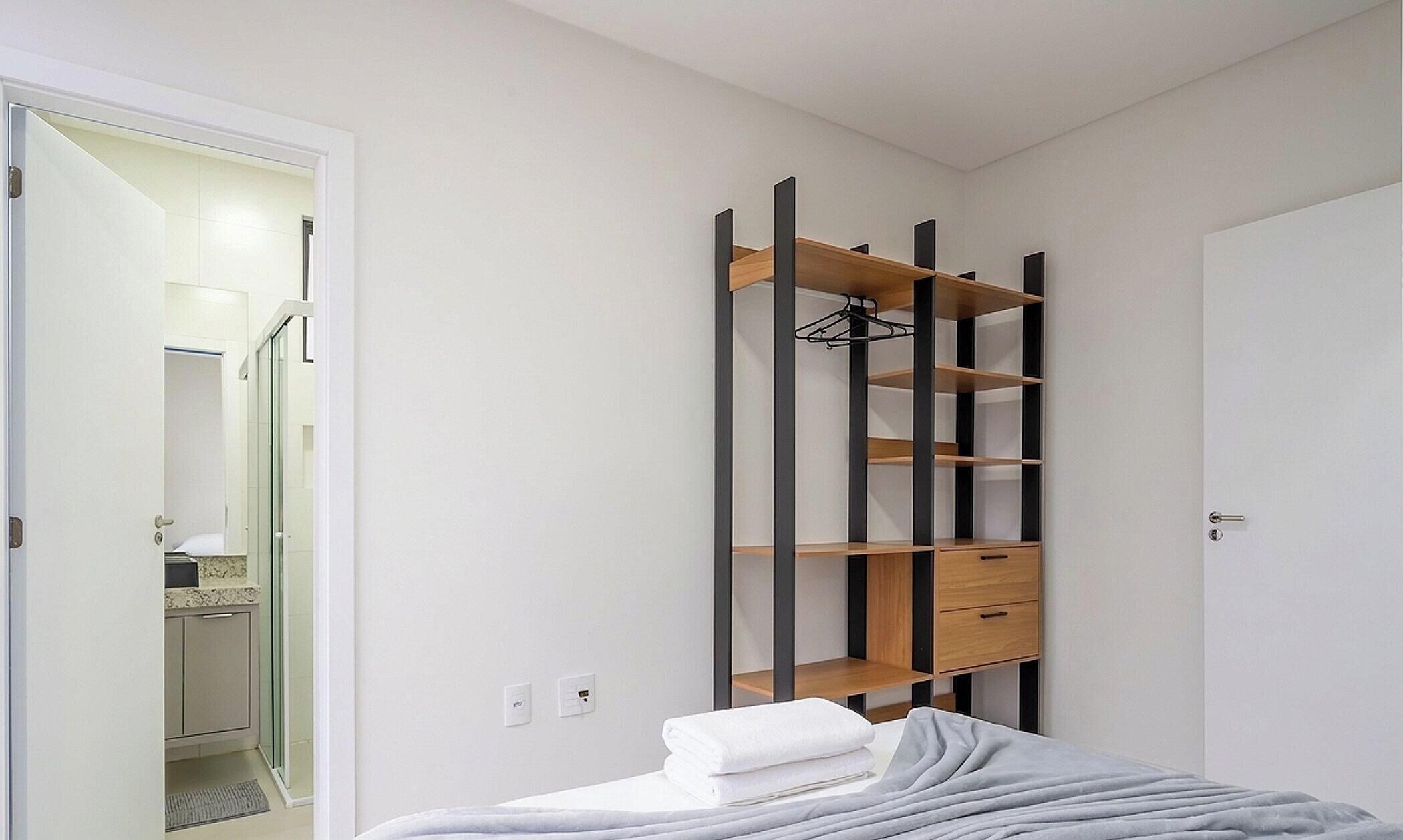 Quarto com cama, guarda-roupa com prateleiras, banheiro com chuveiro e porta para outro cômodo.