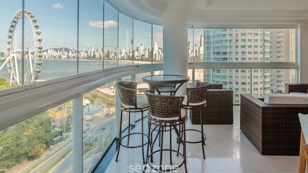 Varanda com mobiliário de vime e guarda-corpo de vidro, com vista para a paisagem urbana e uma roda gigante.