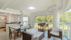 Ampla sala de estar com mesa de jantar e móveis de vime, cozinha com armários brancos e janelas grandes com vista para a floresta.