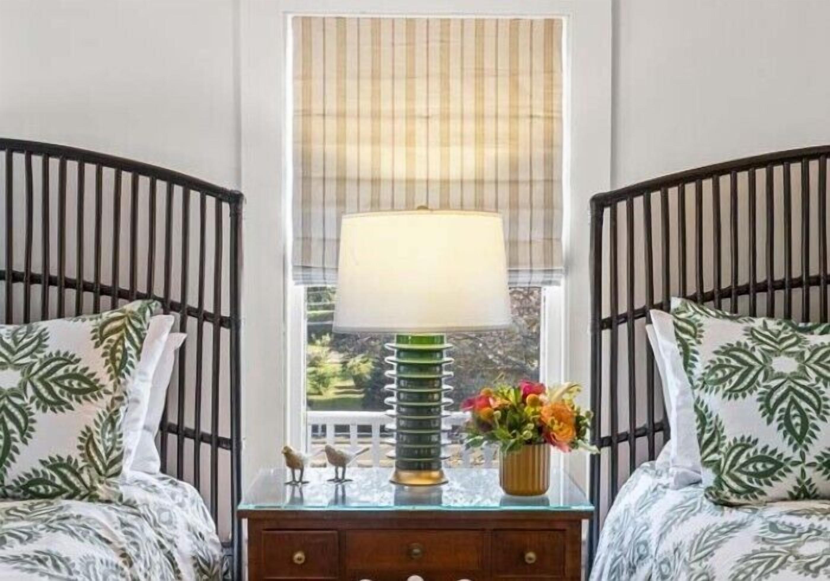 Quarto com duas camas, uma mesa de cabeceira com abajur, uma janela com persianas e vista para o verde lá fora.