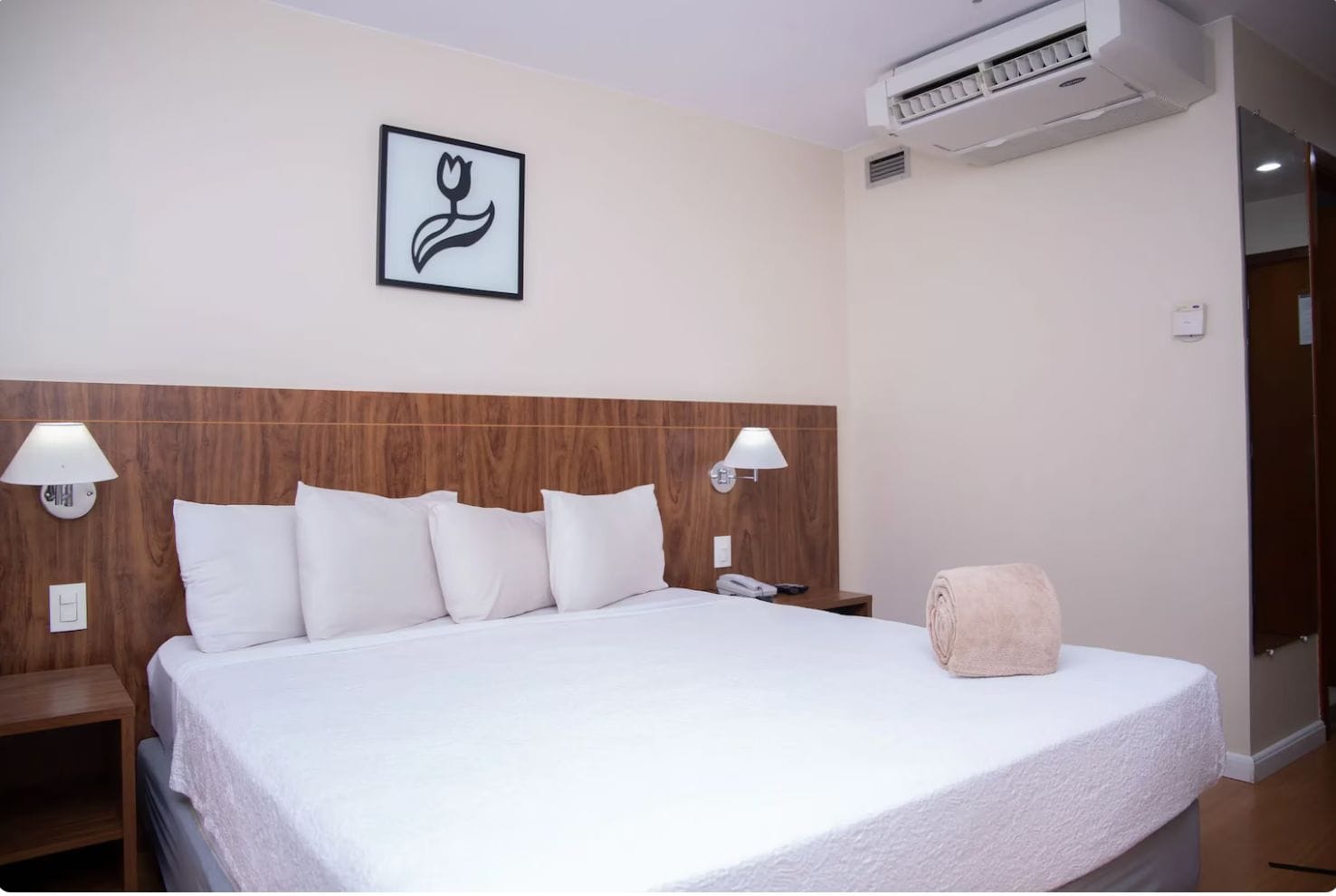 Quarto de hotel com cabeceira de madeira, cama com roupa de cama branca, duas luminárias fixadas na parede, um quadro emoldurado, um aparelho de ar condicionado e uma toalha enrolada na cama.