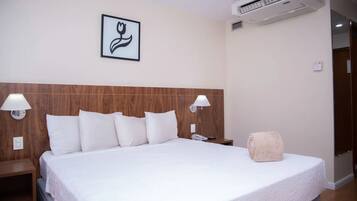Quarto de hotel com cabeceira de madeira, cama com roupa de cama branca, duas luminárias fixadas na parede, um quadro emoldurado, um aparelho de ar condicionado e uma toalha enrolada na cama.