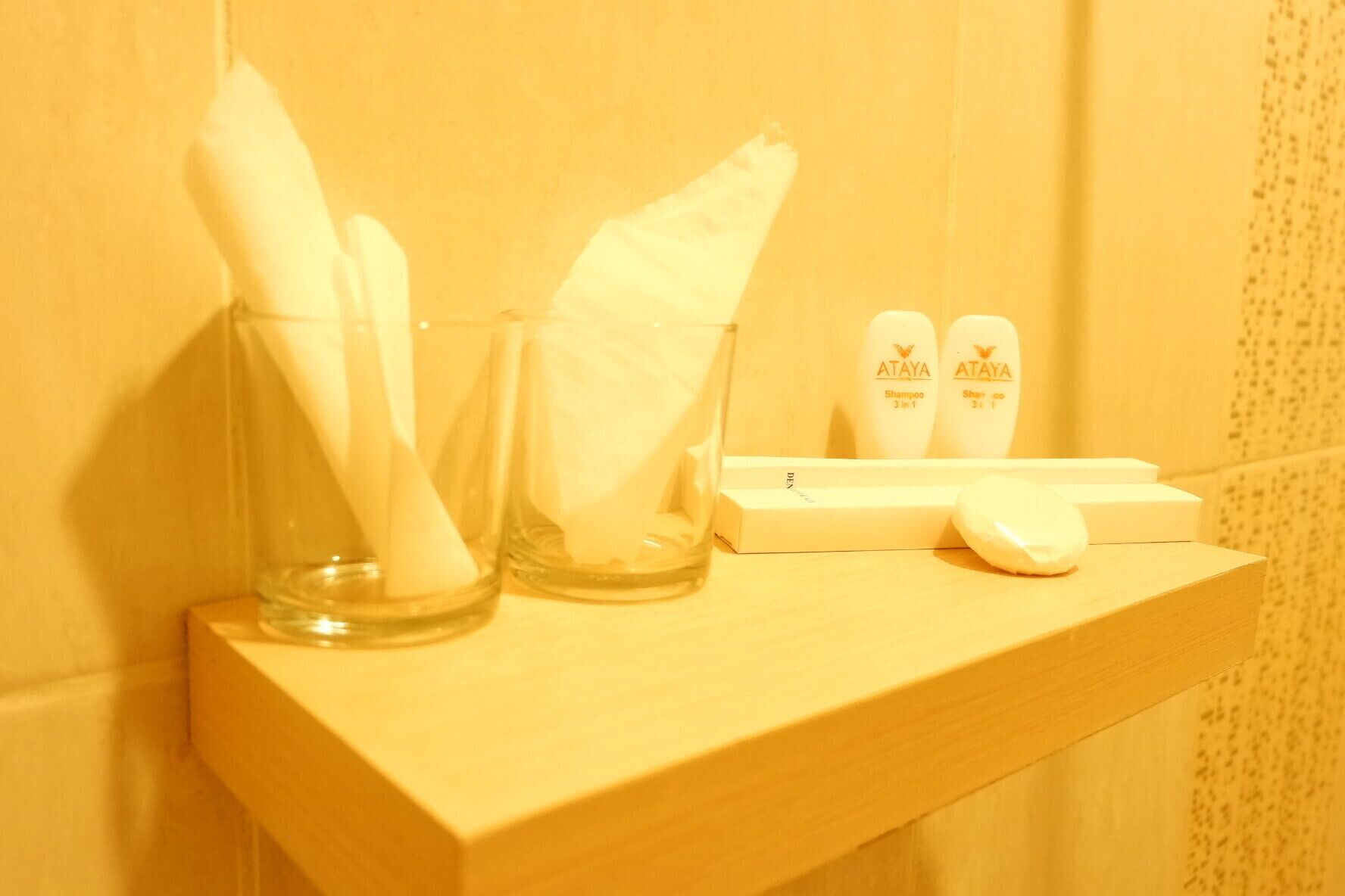 Uma prateleira no banheiro com um copo, duas toalhas brancas e duas garrafas de shampoo Ataya.