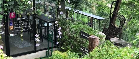 Um café com paredes de vidro, rodeado por vegetação exuberante e um banco de madeira.