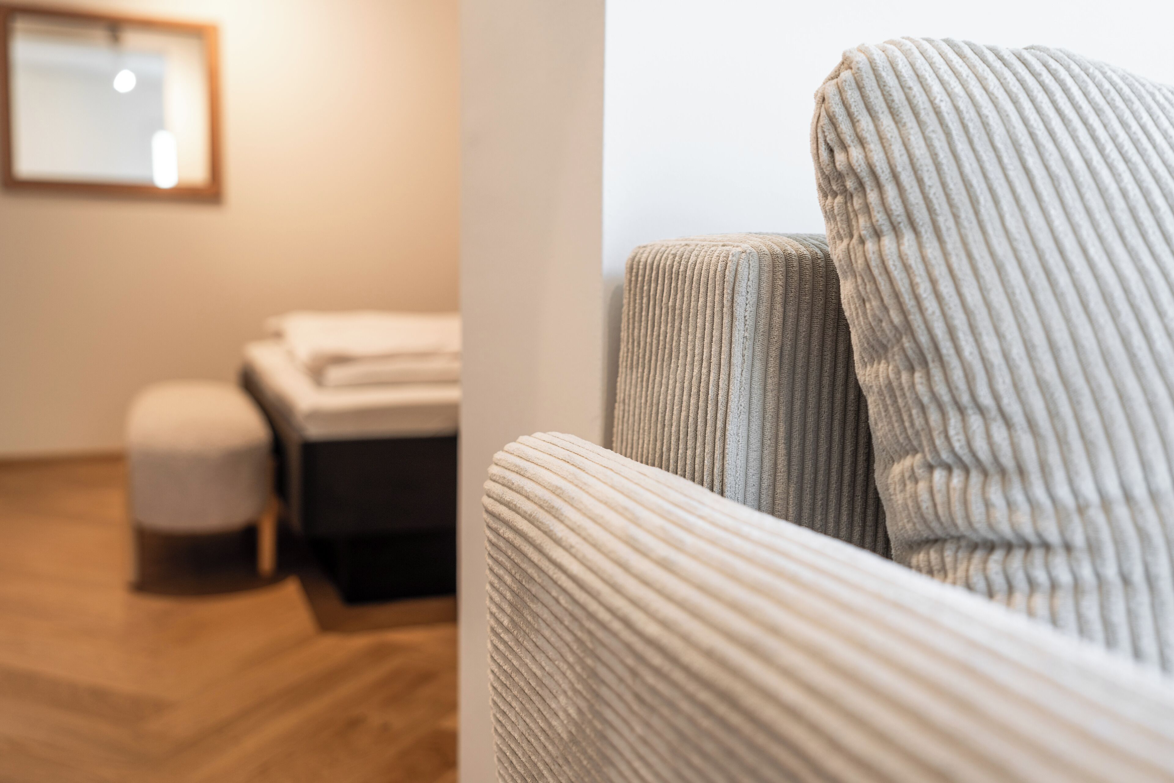 Um close-up de um sofá texturizado em tom claro num quarto moderno e minimalista, com chão de madeira e um espelho emoldurado na parede.