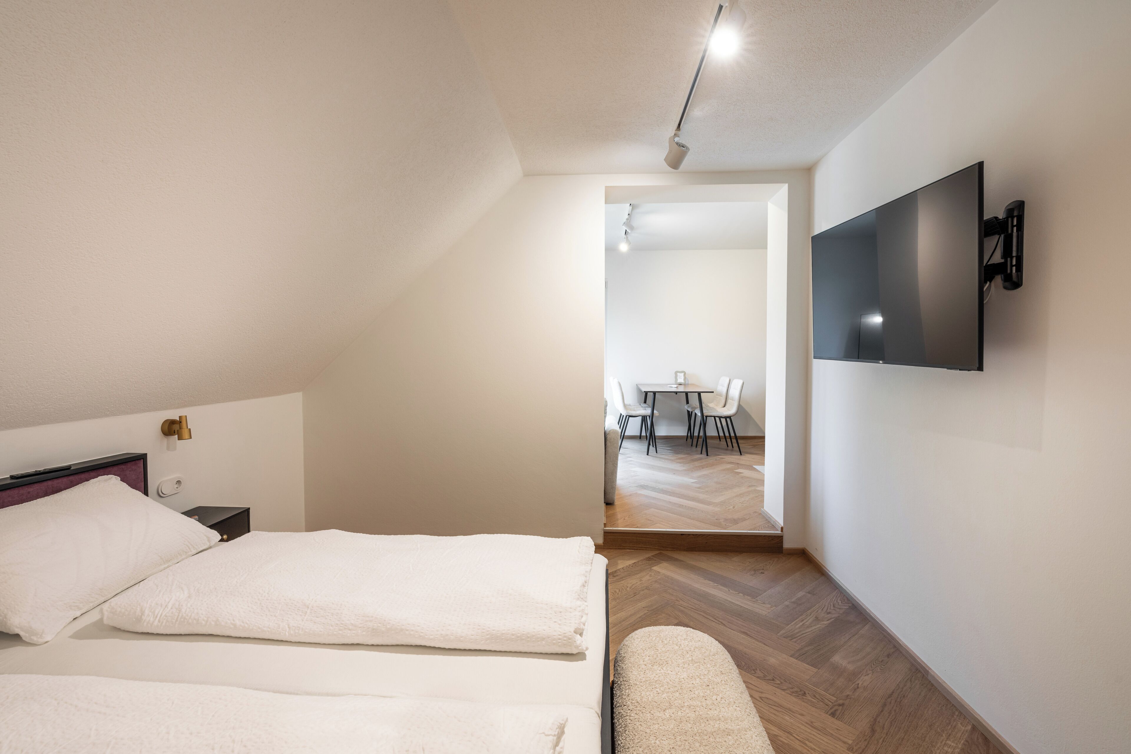 Quarto de hotel moderno com uma cama, uma televisão fixada na parede e uma pequena mesa com cadeiras ao fundo.