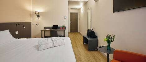 Quarto de hotel com cama, escrivaninha, cadeira, mesa pequena com um vaso de flores, sofá e televisão fixada na parede.