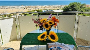 Varanda com mesa posta para dois, apresentando um vaso com girassóis e toalha de mesa verde. A vista inclui uma praia e o mar.