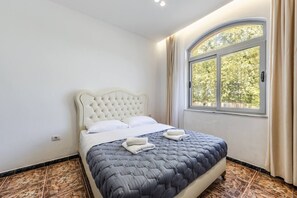 Cama bem arrumada com cabeceira estofada e colcha acolchoada, janela com cortinas e piso estampado.